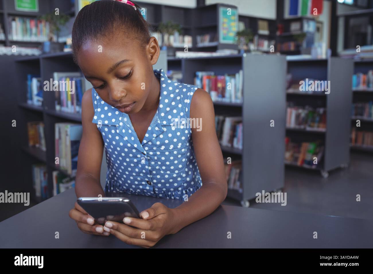 African American girl child using smartphone at library table, among bookshelves and book cart ...