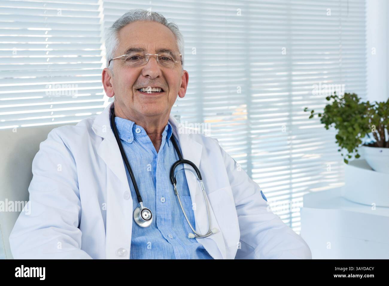 Senior doctor sitting in medical office by window blinds, wearing lab coat, stethoscope, copy ...