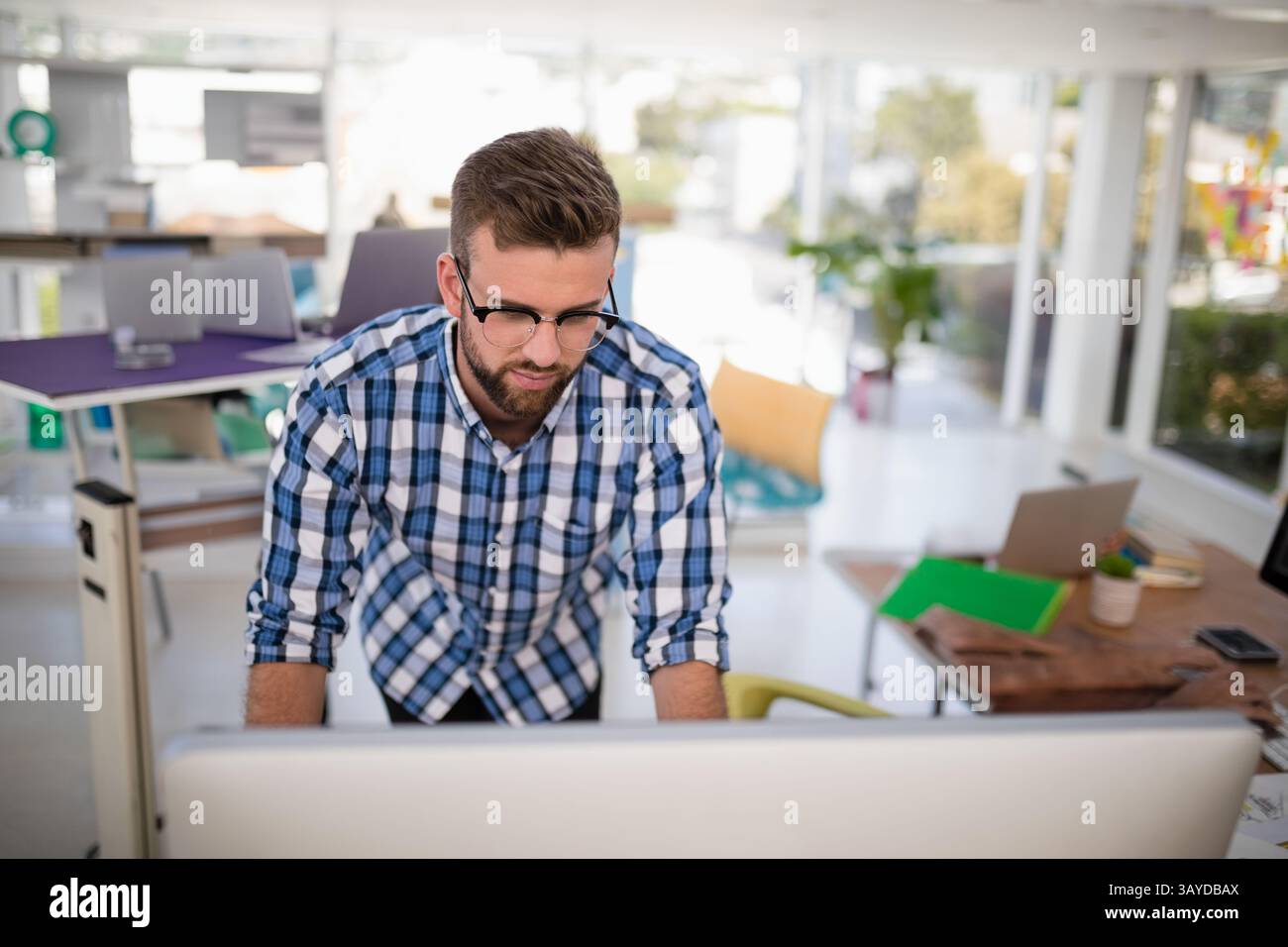 Man leaning over large desktop monitor in modern open-plan office, with ...