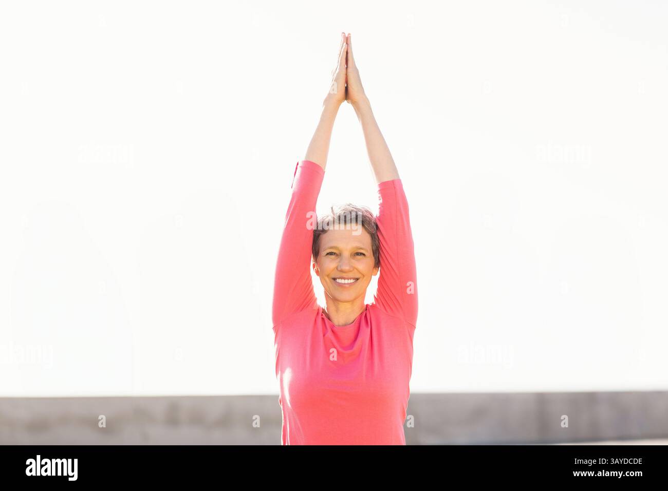 Woman stretching in front of white wall outdoors, holding arms overhead ...