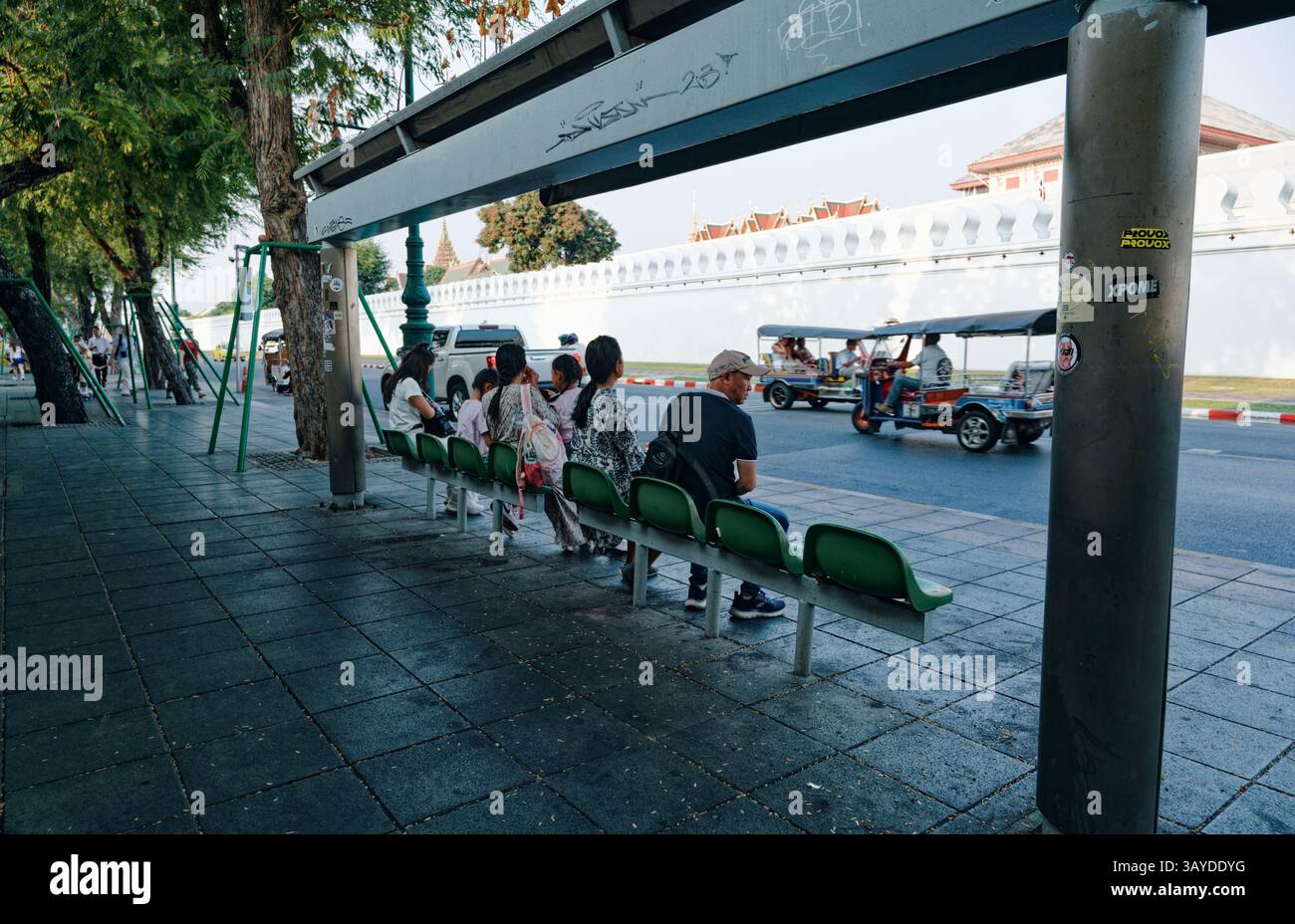 Locals and tourists sit patiently at a bus stop as colorful tuk-tuks ...