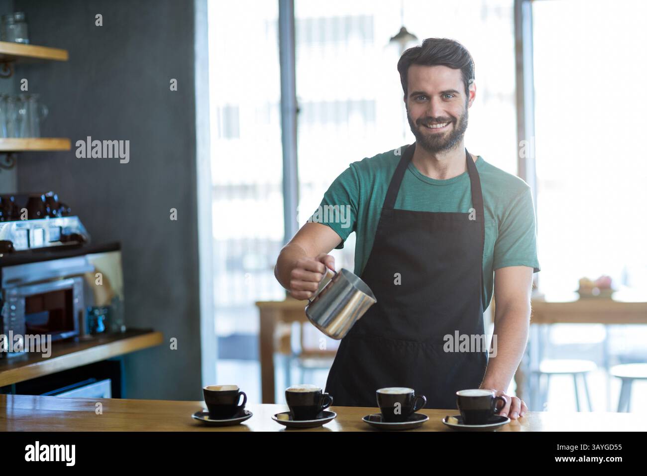 Young male barista pouring milk from steel pitcher into coffee cups on ...