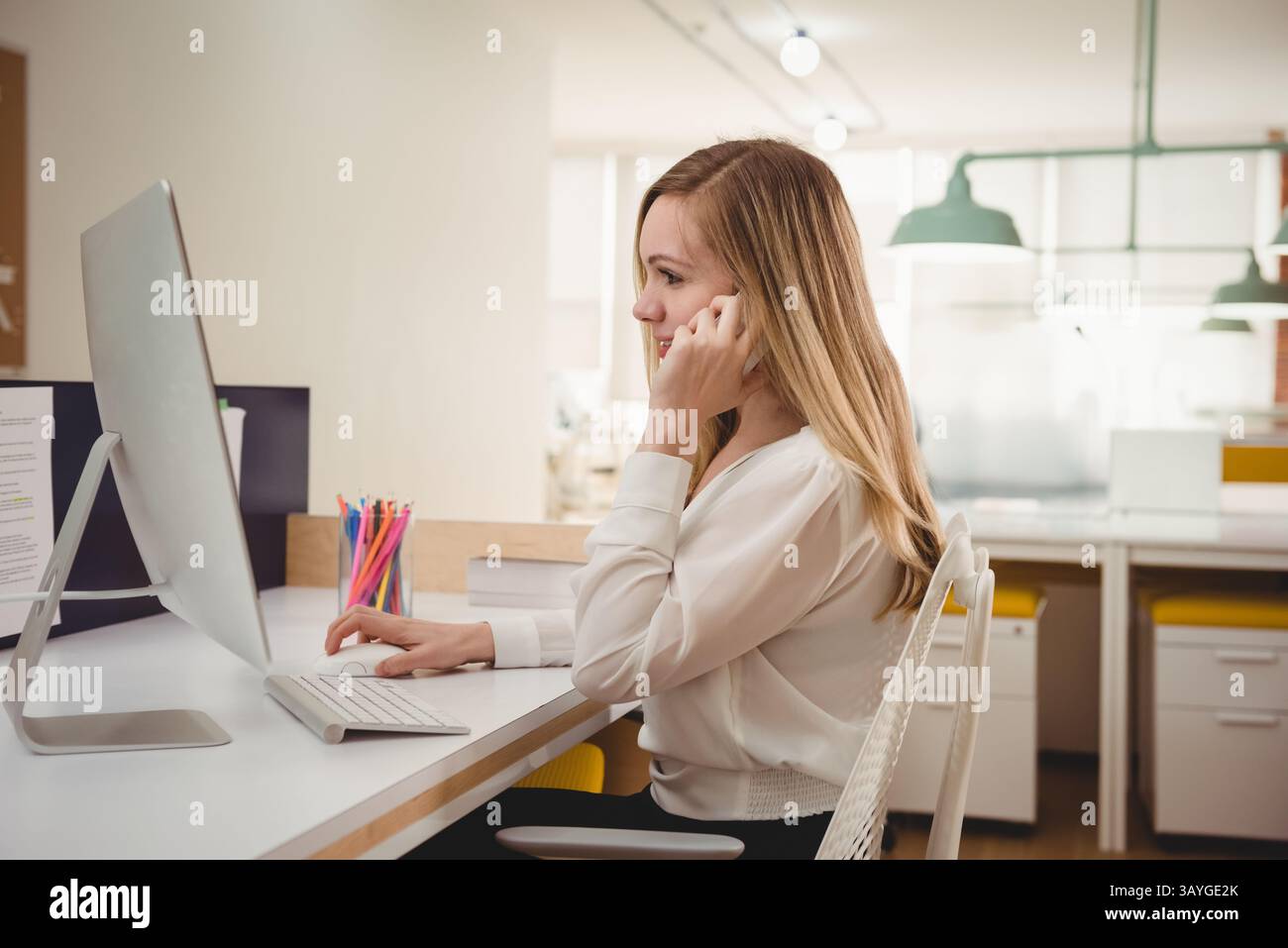 Woman talking on smartphone while typing on keyboard in open-plan ...