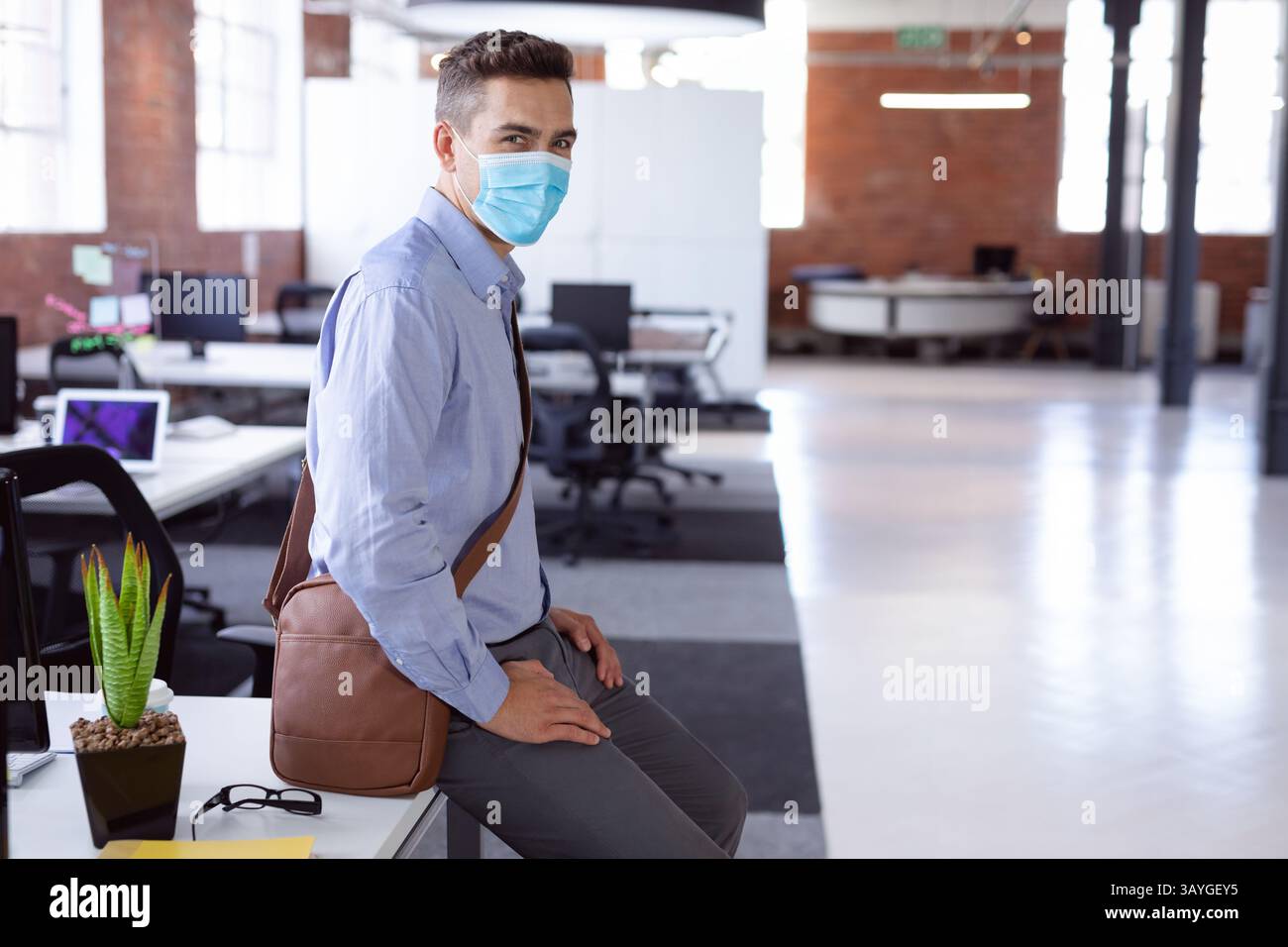 Young man sitting on desk edge in industrial open-plan office, wearing ...