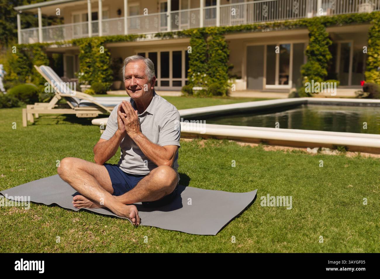 Senior man sitting cross-legged on gray yoga mat in backyard ...