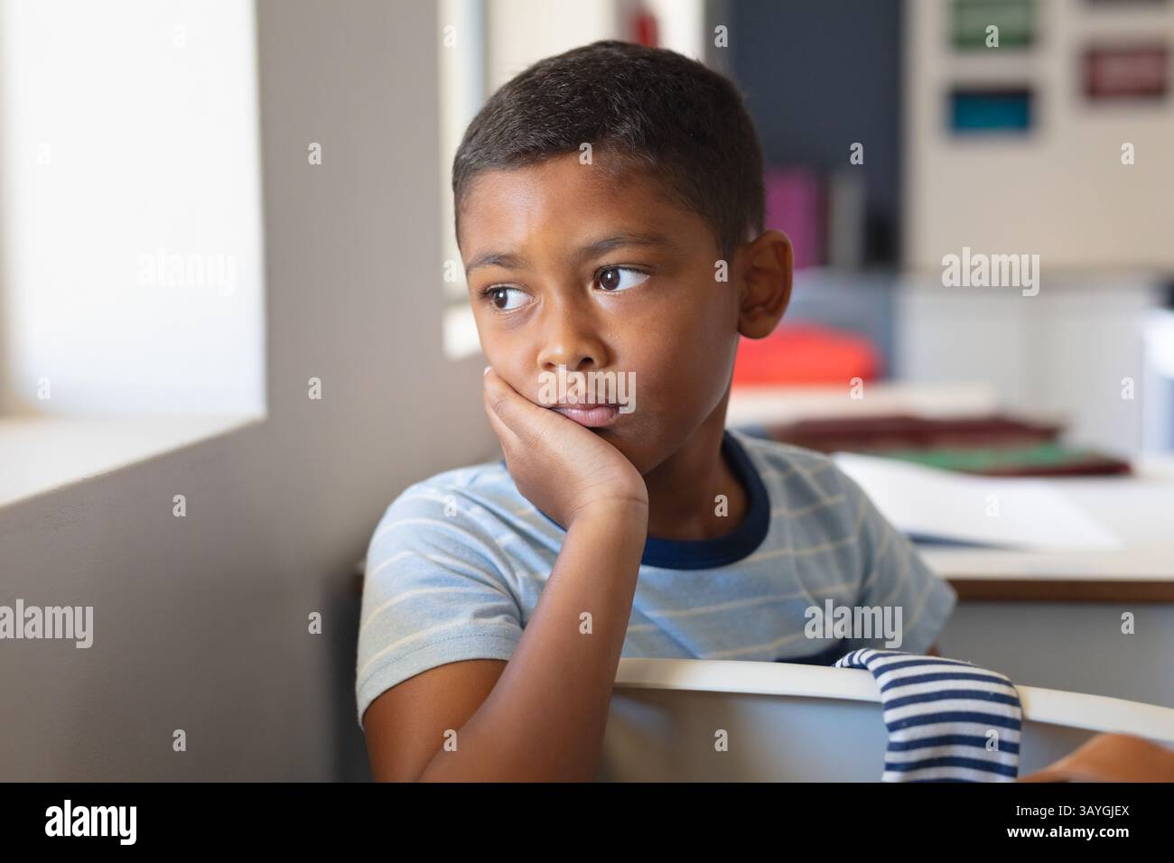7-year-old boy resting cheek on hand and gazing outside at classroom ...