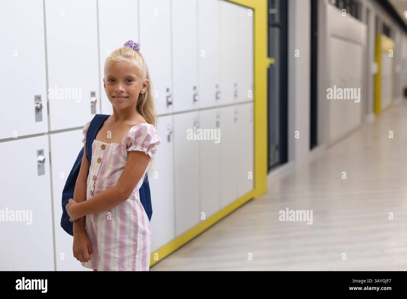 Child girl standing in school corridor with yellow lockers, carrying navy blue backpack, copy ...