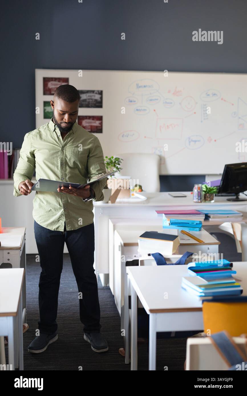 African American male teacher reading folder in modern classroom, with ...