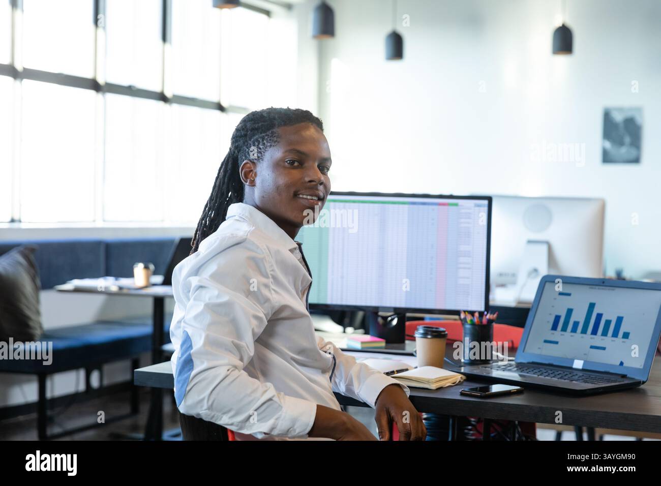 African American man working at desk in modern open-plan office, with laptop, monitor, coffee ...