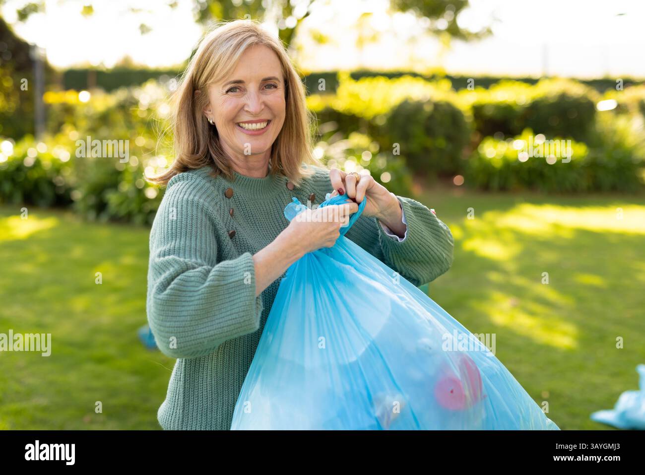 Woman in her fifties tying off blue plastic garbage bag on backyard ...