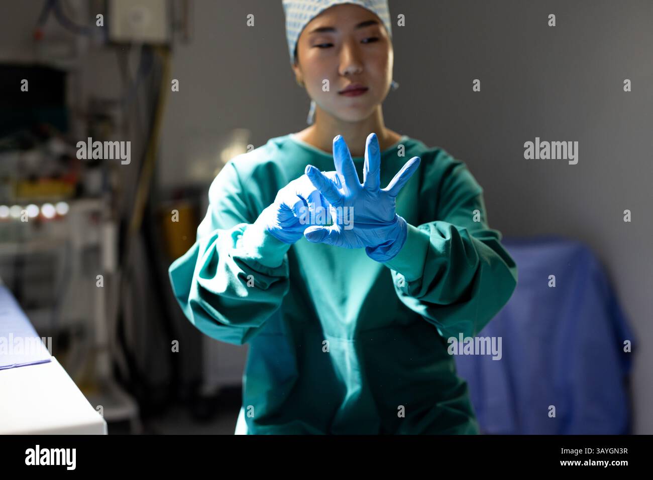 Young Korean woman surgeon pulling on blue gloves in dim operating room ...
