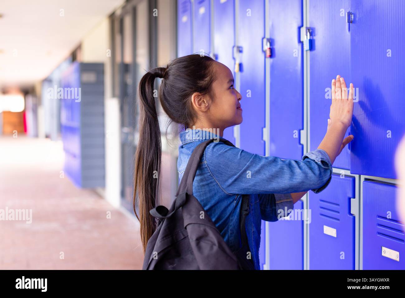 Asian girl touching blue lockers in school hallway, wearing denim ...