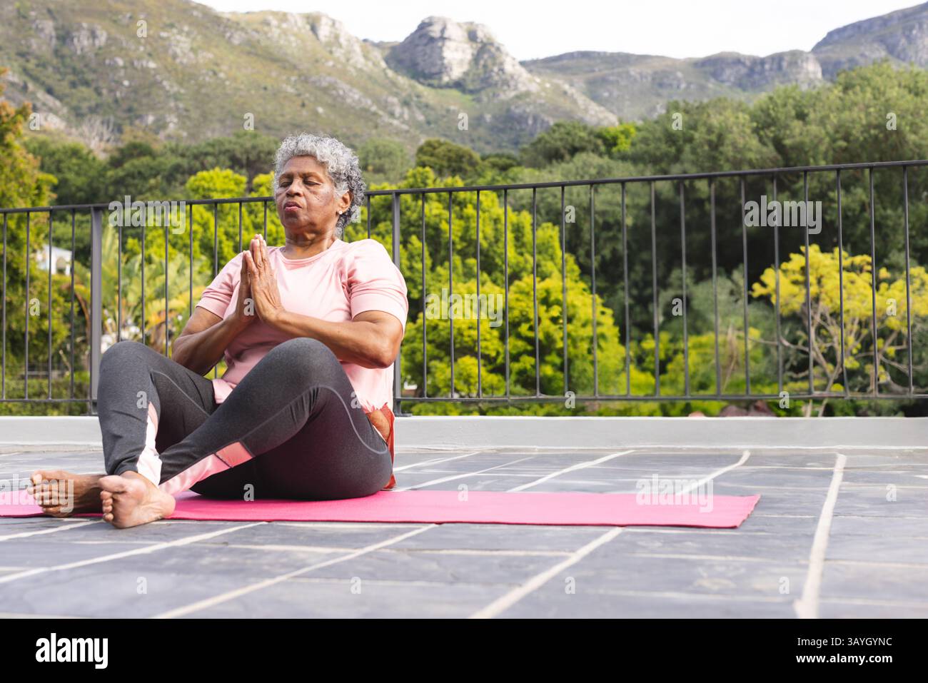Senior African American woman practicing prayer pose on outdoor terrace, using pink yoga mat ...