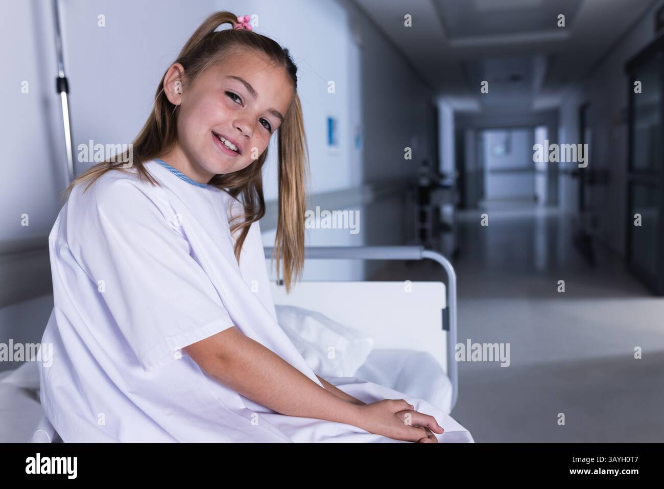 Female child sitting on hospital bed in hospital corridor, wearing gown with metal rails and ...