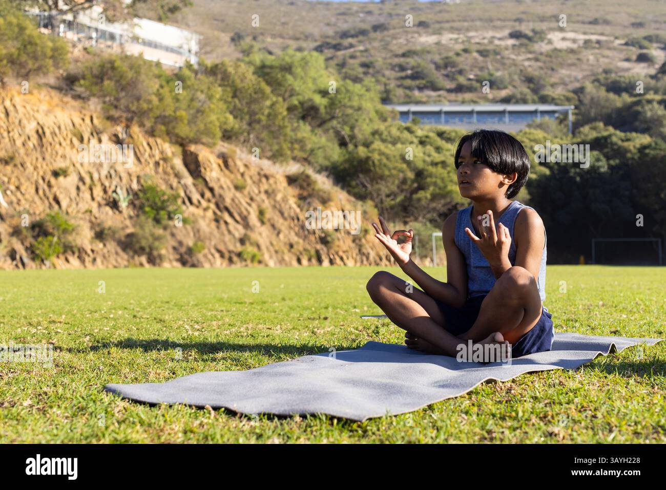 Multiracial boy sitting cross-legged on gray yoga mat in park, holding ...