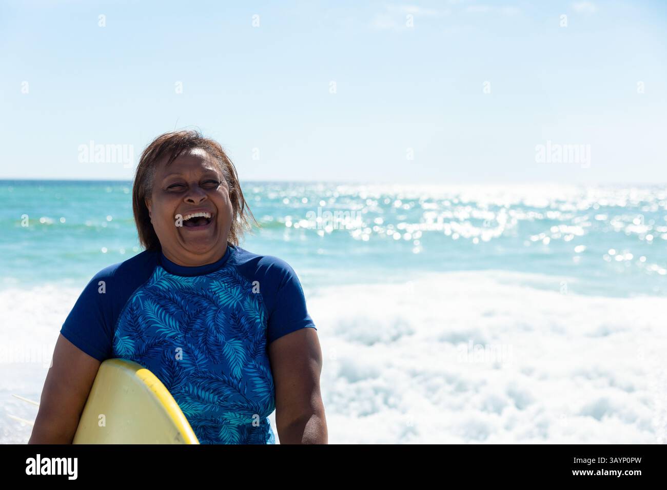 Senior woman standing at shoreline wearing blue rash guard and holding ...