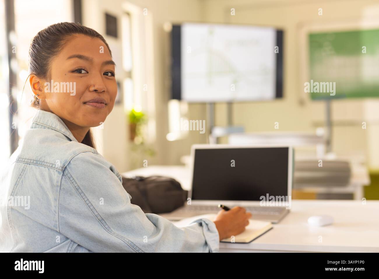 Asian female student writing in notebook at desk in classroom with ...