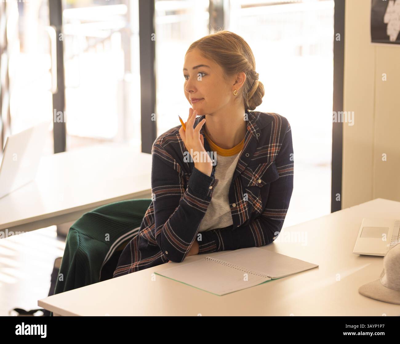 young female student holding pencil and gazing out window in study ...