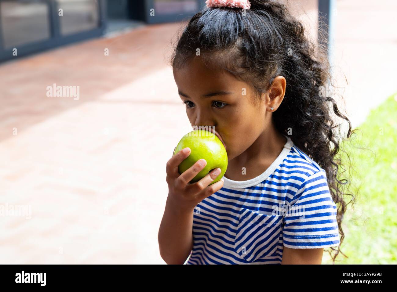 Multiracial girl child biting bright green apple on brick patio at home ...