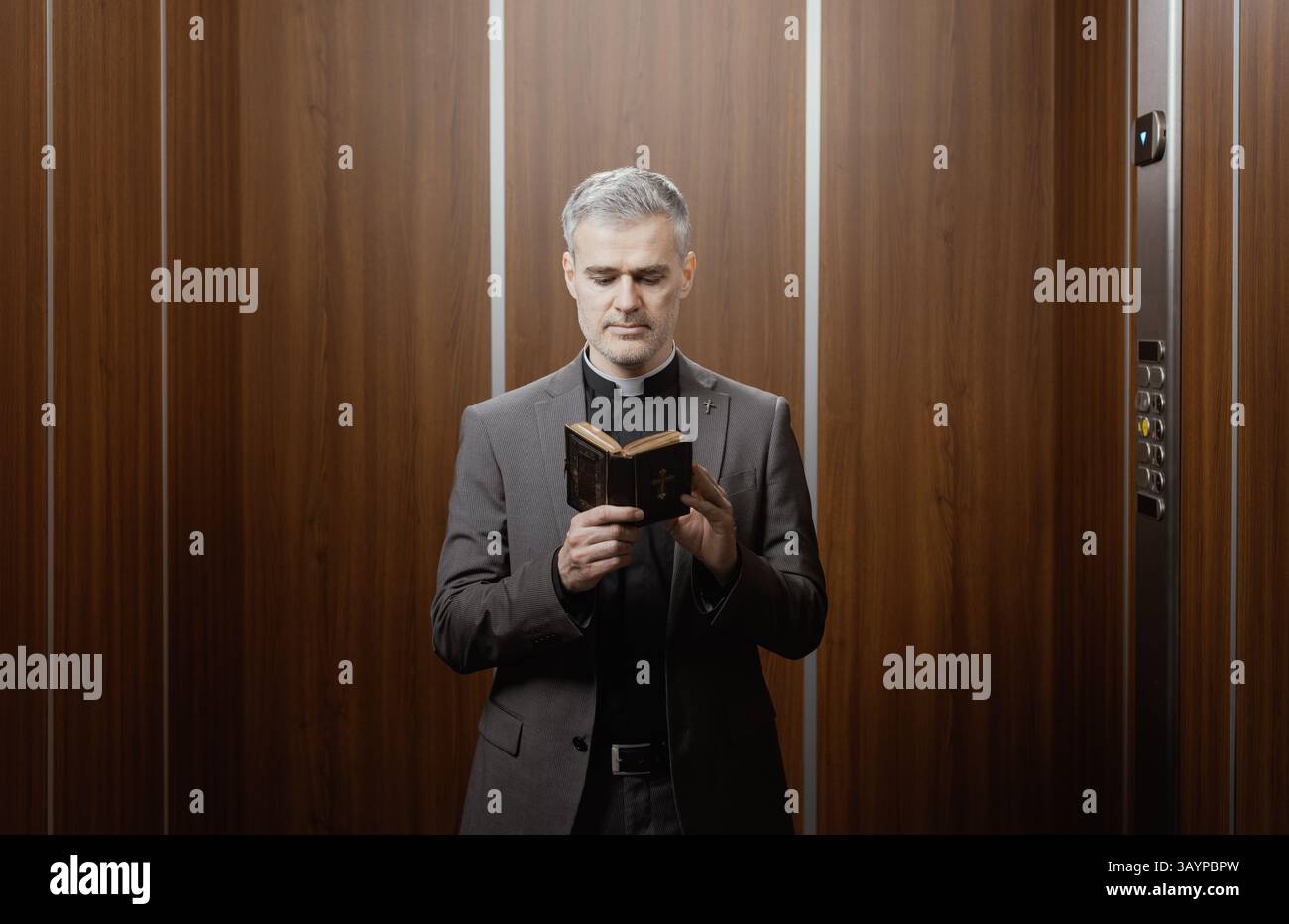 Focused priest standing in an elevator and reading prayers in a book ...