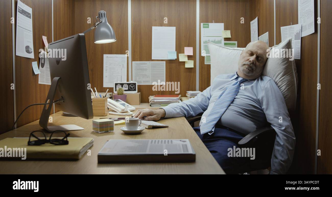 Lazy senior office worker sleeping at his desk in a tiny office, he is ...