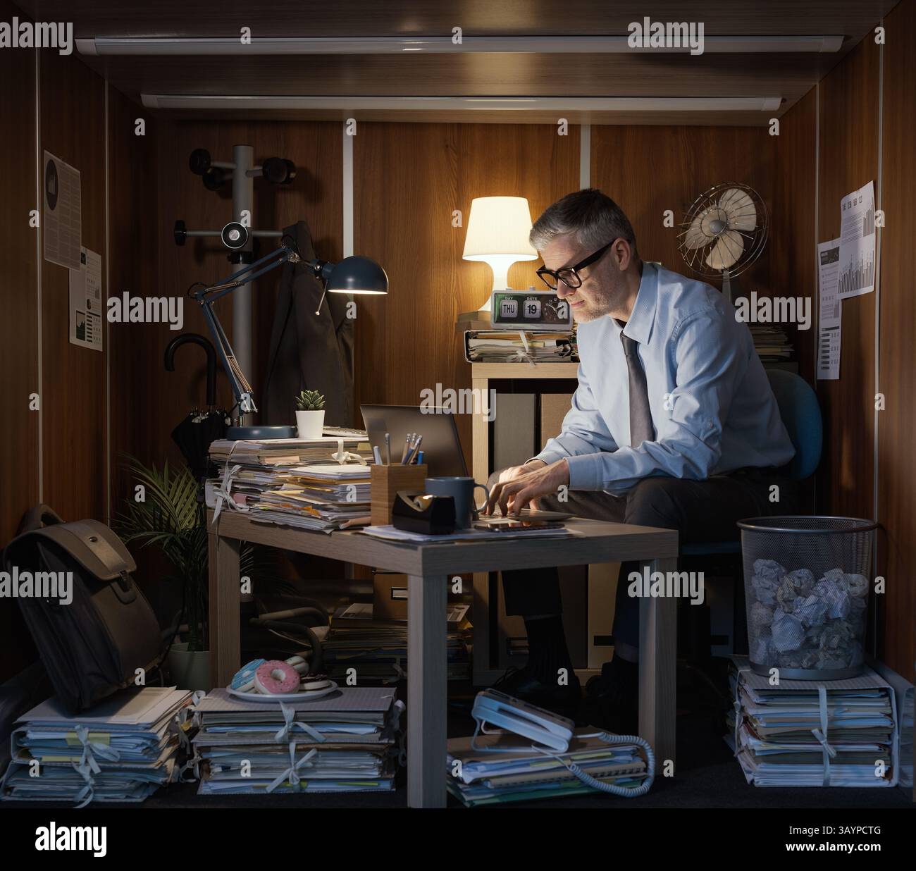 Workaholic businessman sitting at his desk and working late at night in a small office, he is ...