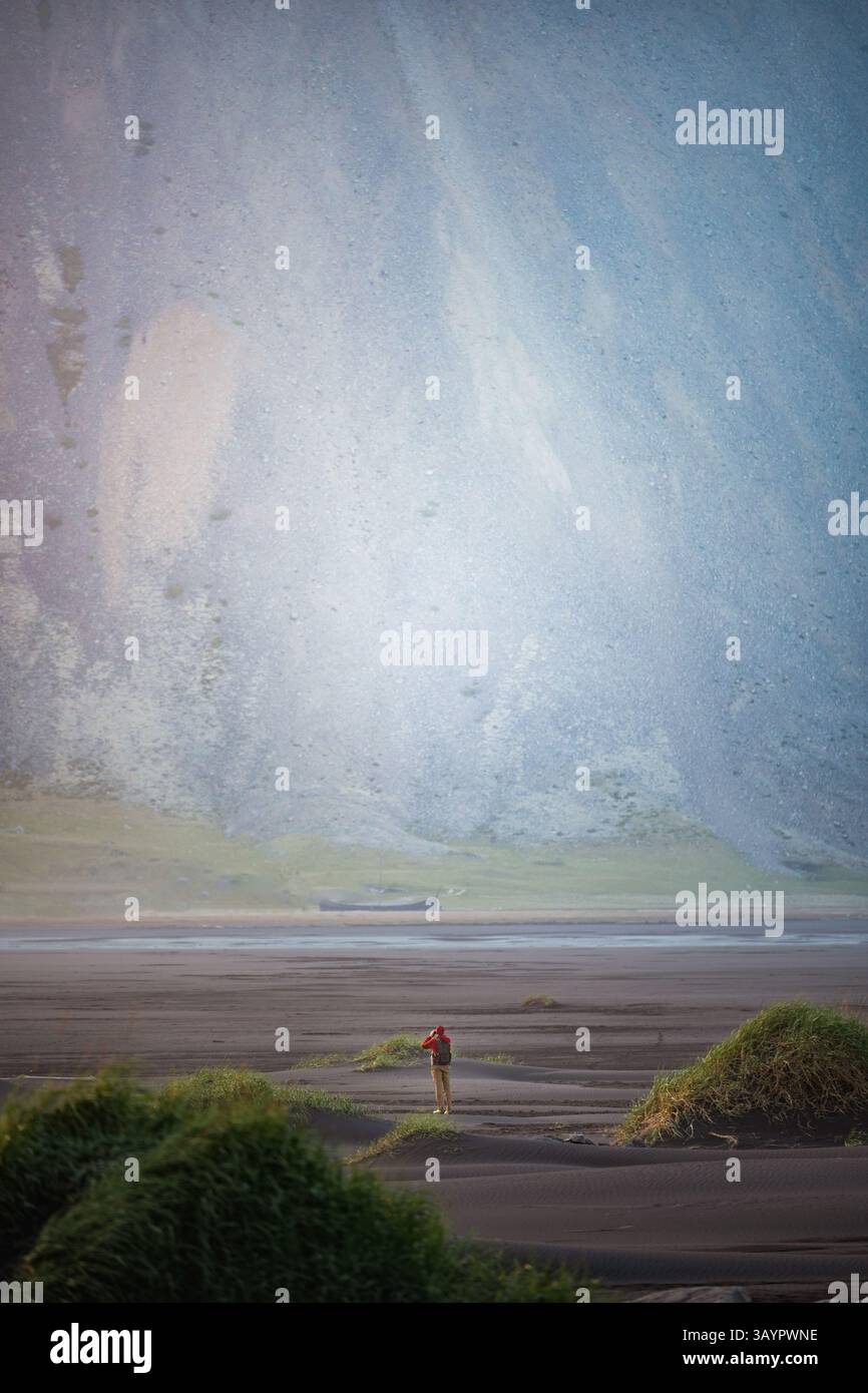 Lone hiker with red jacket walks across black sand dunes in front of ...