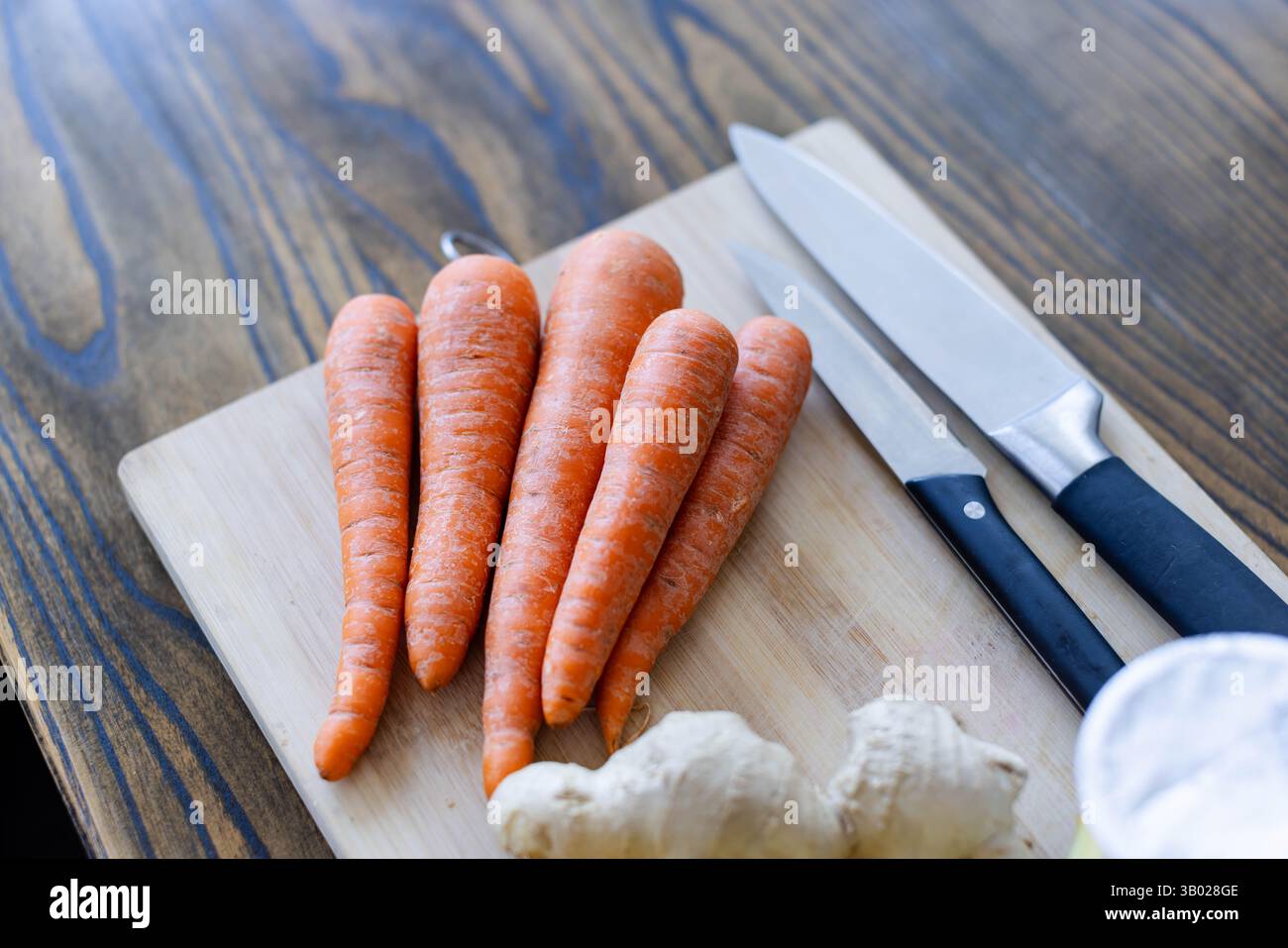 Light cutting board is resting on kitchen table, displaying carrots, knives and ginger root ...