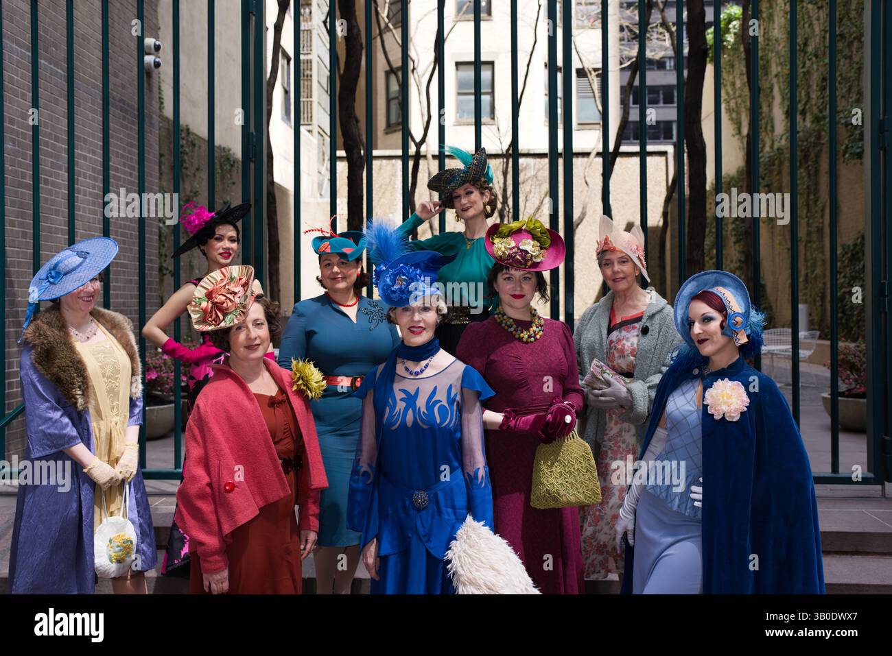 Nine women in vintage-style dresses and hats pose in front of a metal ...