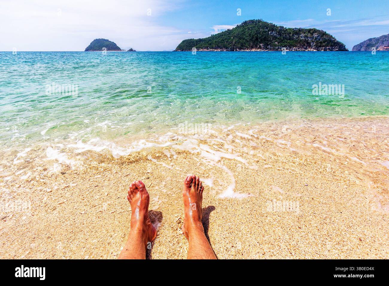 A man’s feet on the white sand of a sandbar with emerald waters and ...