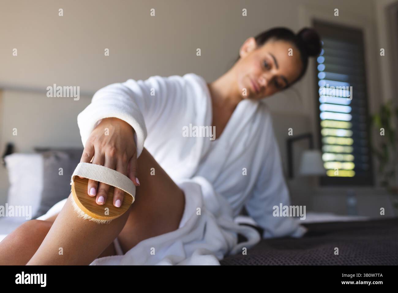 Woman brushing lower leg wearing white bathrobe in bedroom, with wooden ...