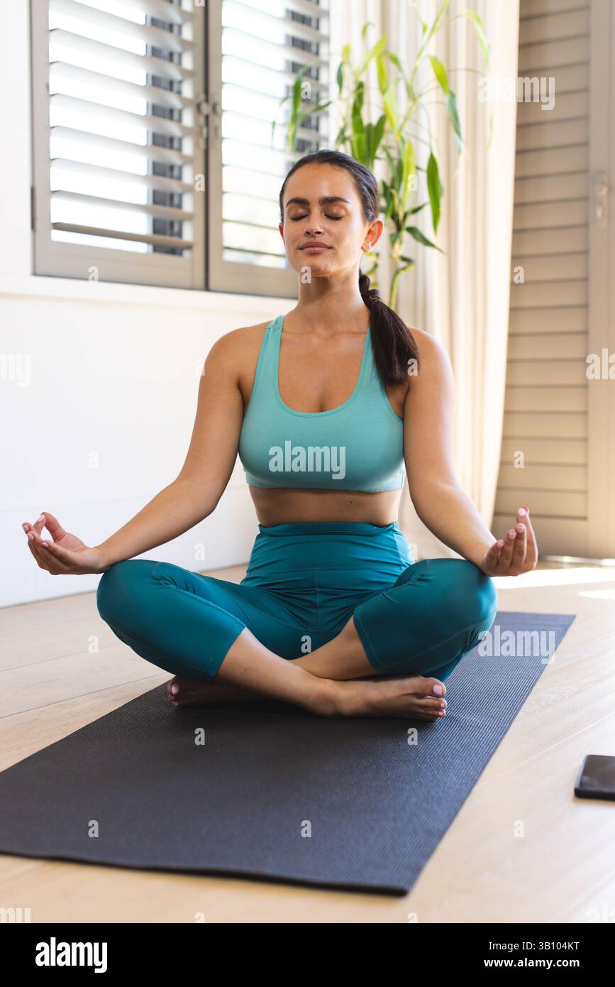 Meditating woman sitting cross-legged in living room, with yoga mat smartphone and plant Stock ...