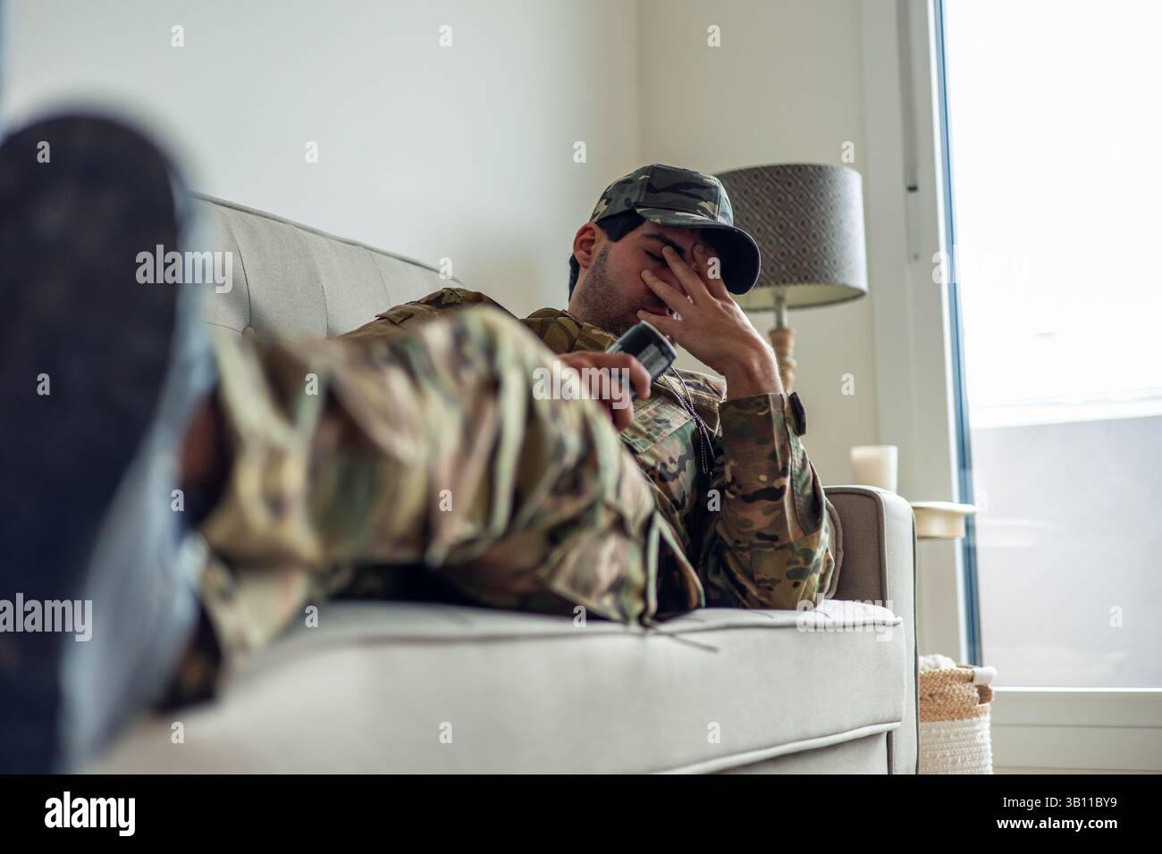 Young soldier in uniform lying on the sofa at home, holding a remote ...