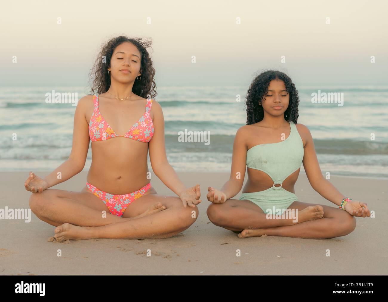 Two women practicing meditation in a serene beach setting. They sit cross-legged on the sand ...