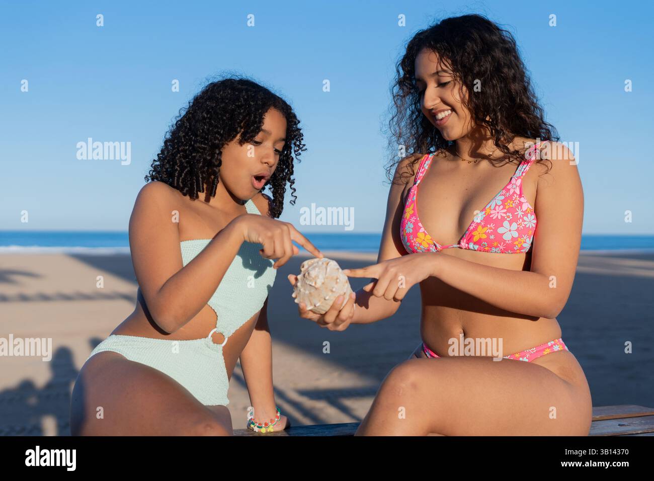Two young women sitting on the beach, happily examining a seashell. The clear blue sky and ocean ...