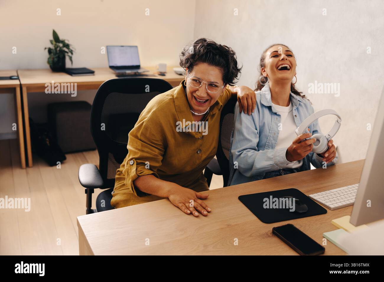 Two coworkers share a joyful moment while seated at a desk in a modern ...