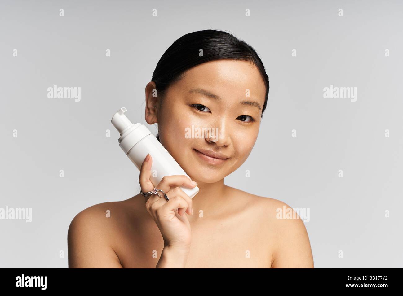 Young asian woman displays skincare product while smiling confidently in minimal studio backdrop ...