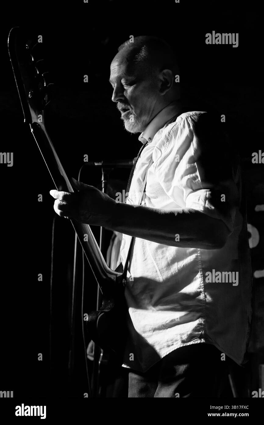 Horace Pantert, lit by a spotlight, plays his bass guitar with focused ...