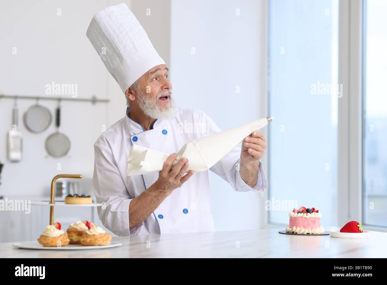 Funny confectioner holding pastry bag at table with desserts in kitchen ...