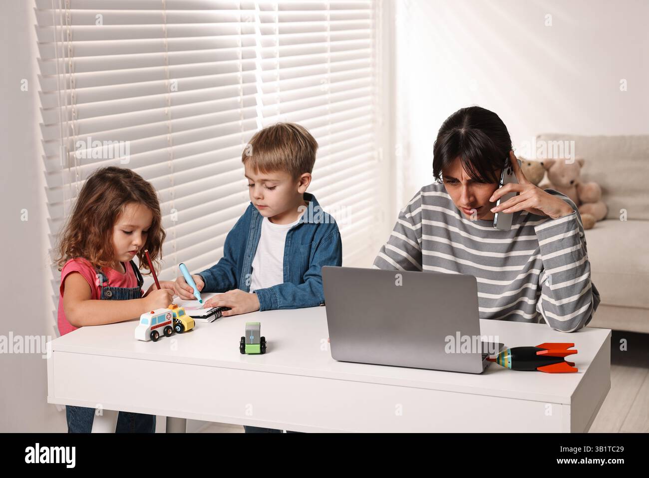 Naughty children drawing while their overwhelmed mother talking on smartphone at table indoors ...