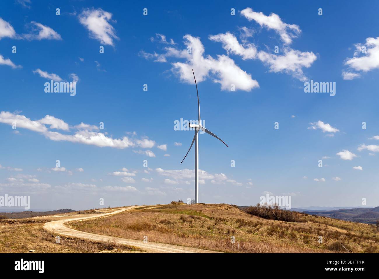 Solitary wind turbine on hilltop with winding dirt road under blue sky ...