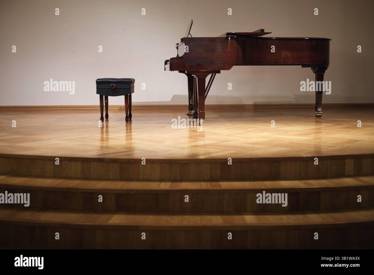 Grand piano and piano bench on empty wooden concert stage against plain ...