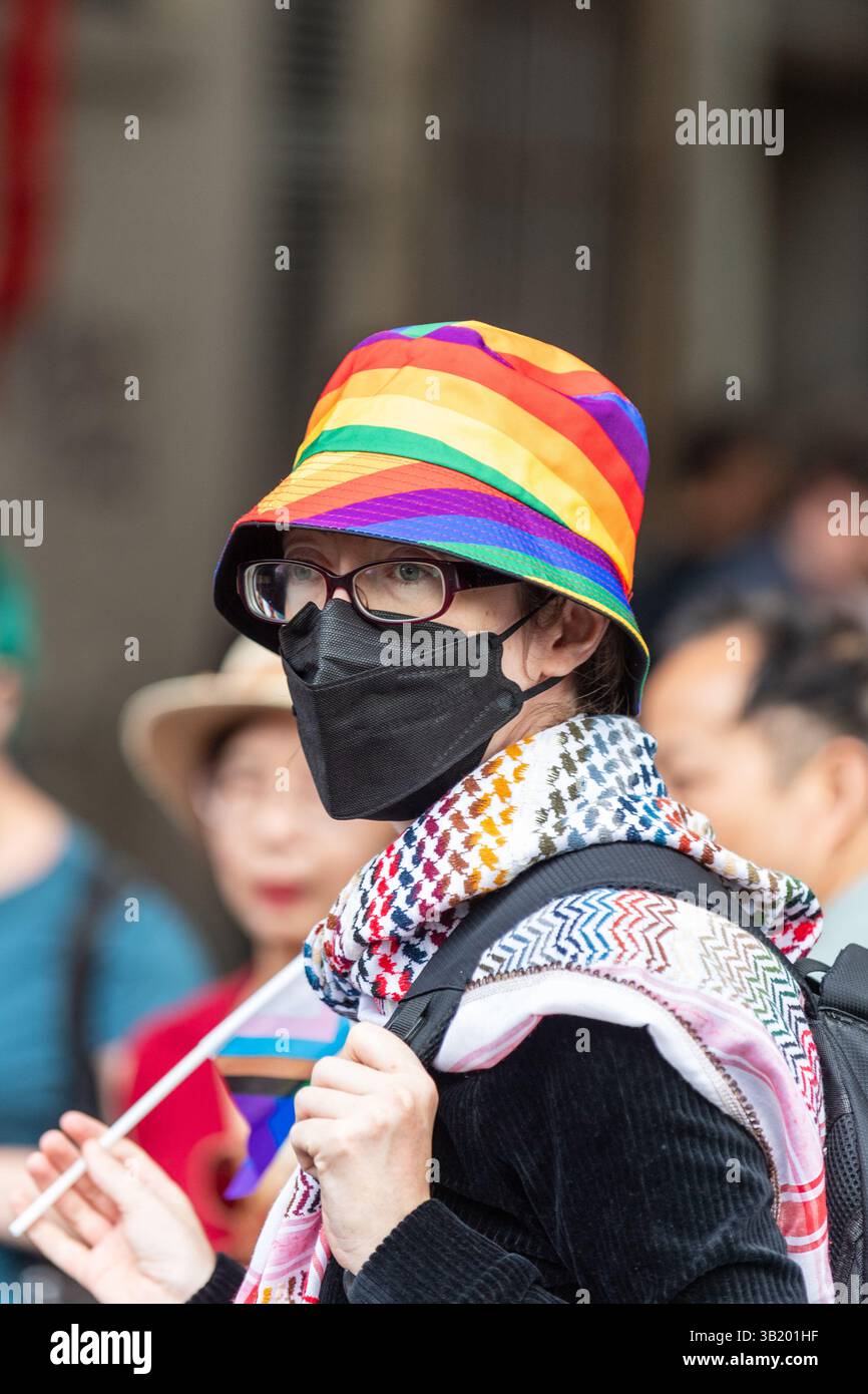 A masked pro-trans protester wearing a rainbow pride hat and keffiyeh ...
