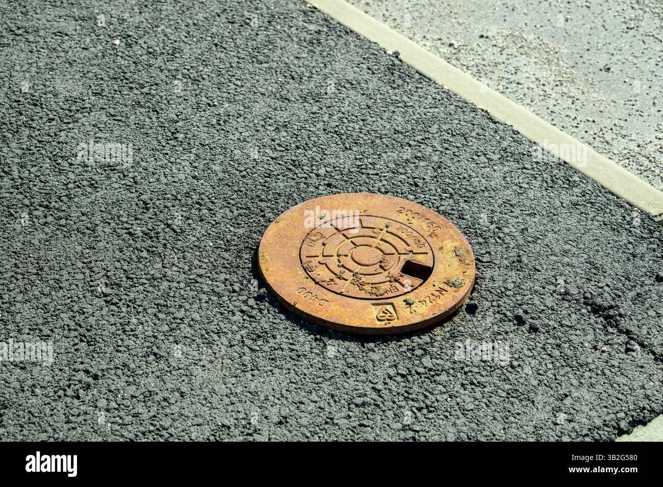 A rusty manhole cover lies embedded in a textured asphalt road surface ...