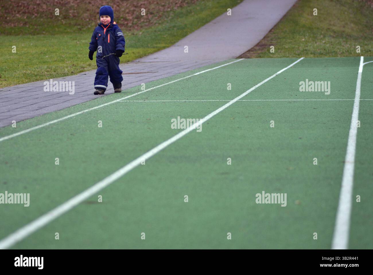 Toddler boy is walking down the footpath next to green running track ...