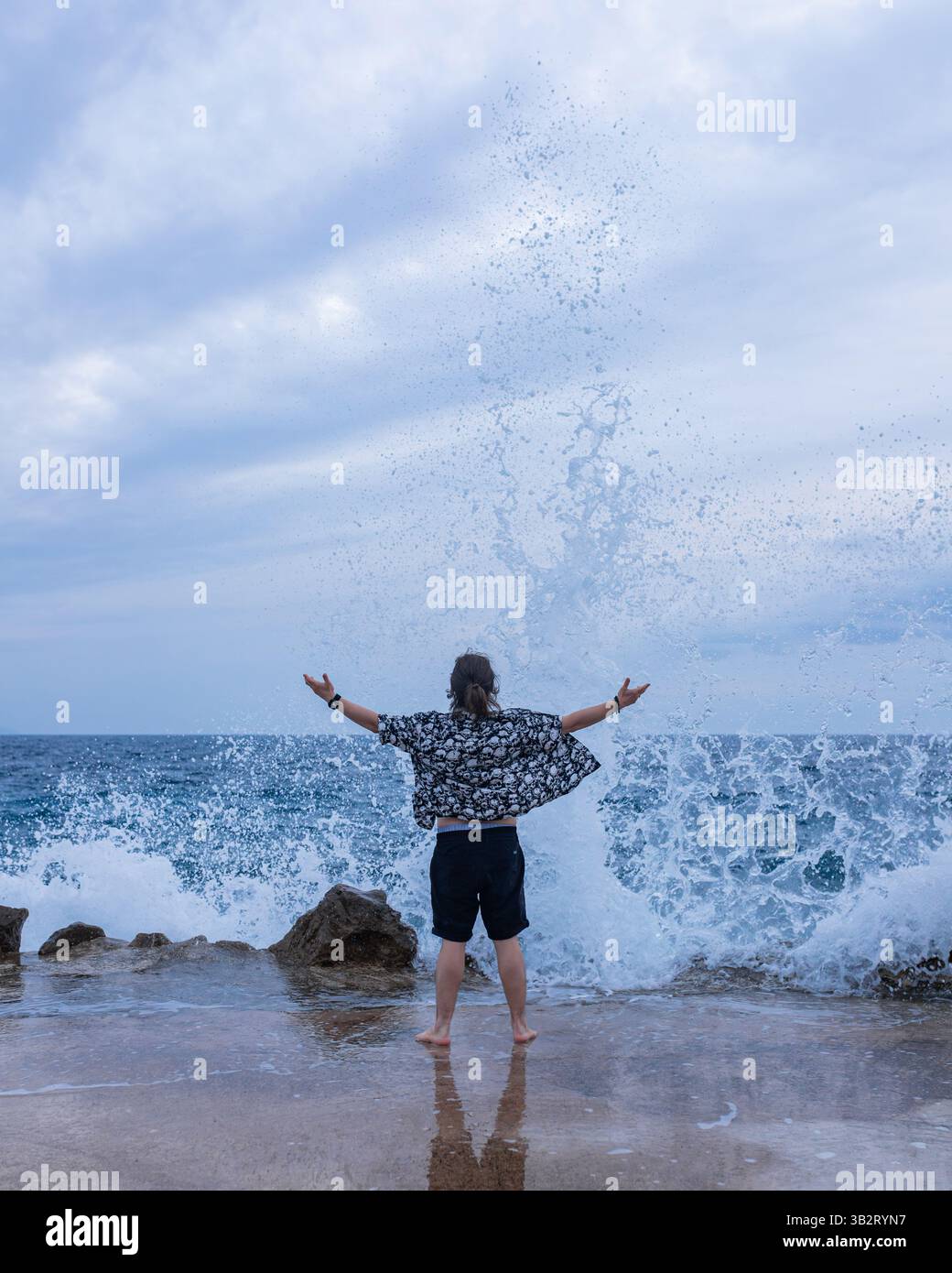 Man stands with arms raised on wet concrete pier facing crashing ocean ...