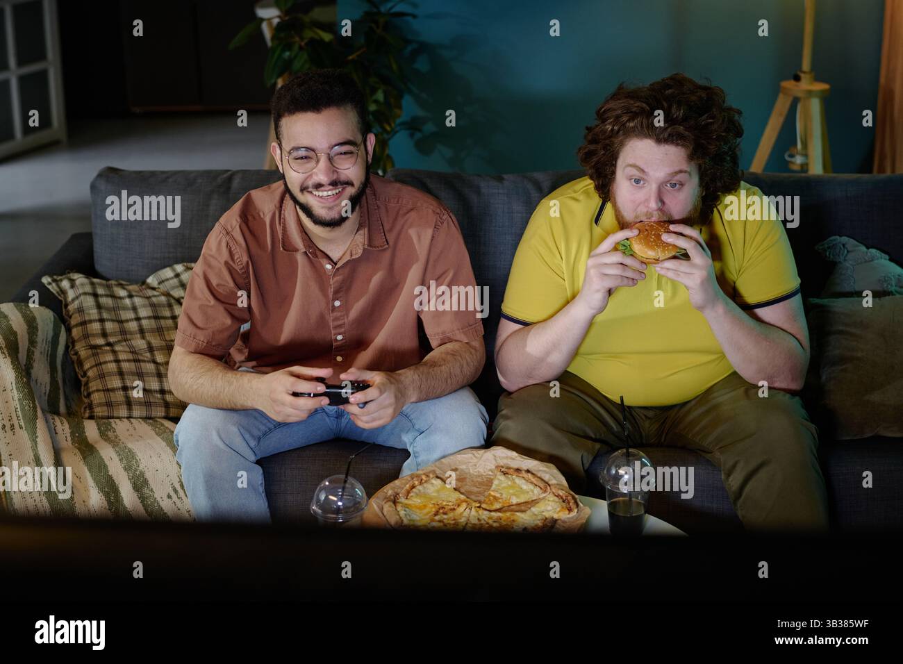 Two friends enjoying video games on couch with snacks and drinks. One ...