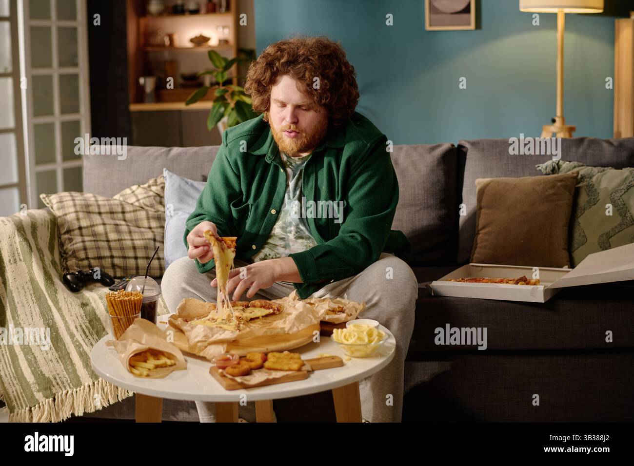 Man with curly hair enjoying pizza and fries while gaming on couch ...