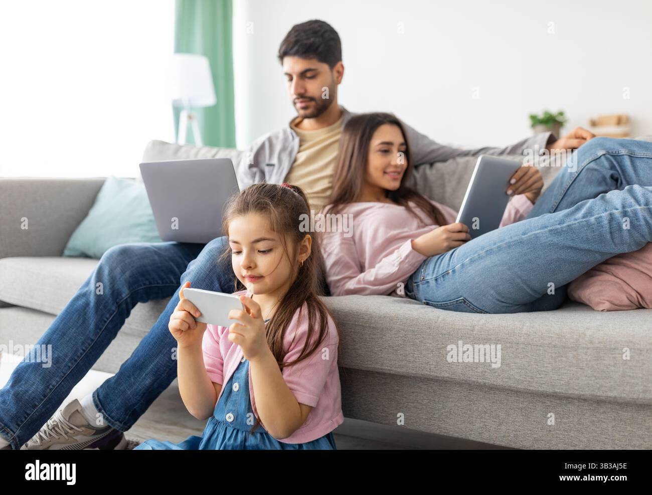 Gadgets addiction. Eastern family using different electronic devices, resting together in living ...