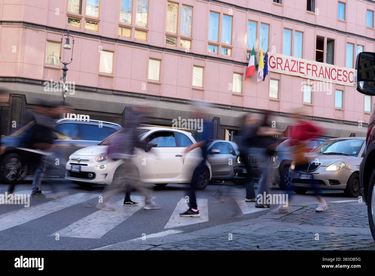 City traffic and pedestrians flow in front of a banner reading "Thank ...